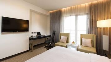 Quarto de hotel com cama, secretária com computador, duas cadeiras, televisão e janela com cortinas.