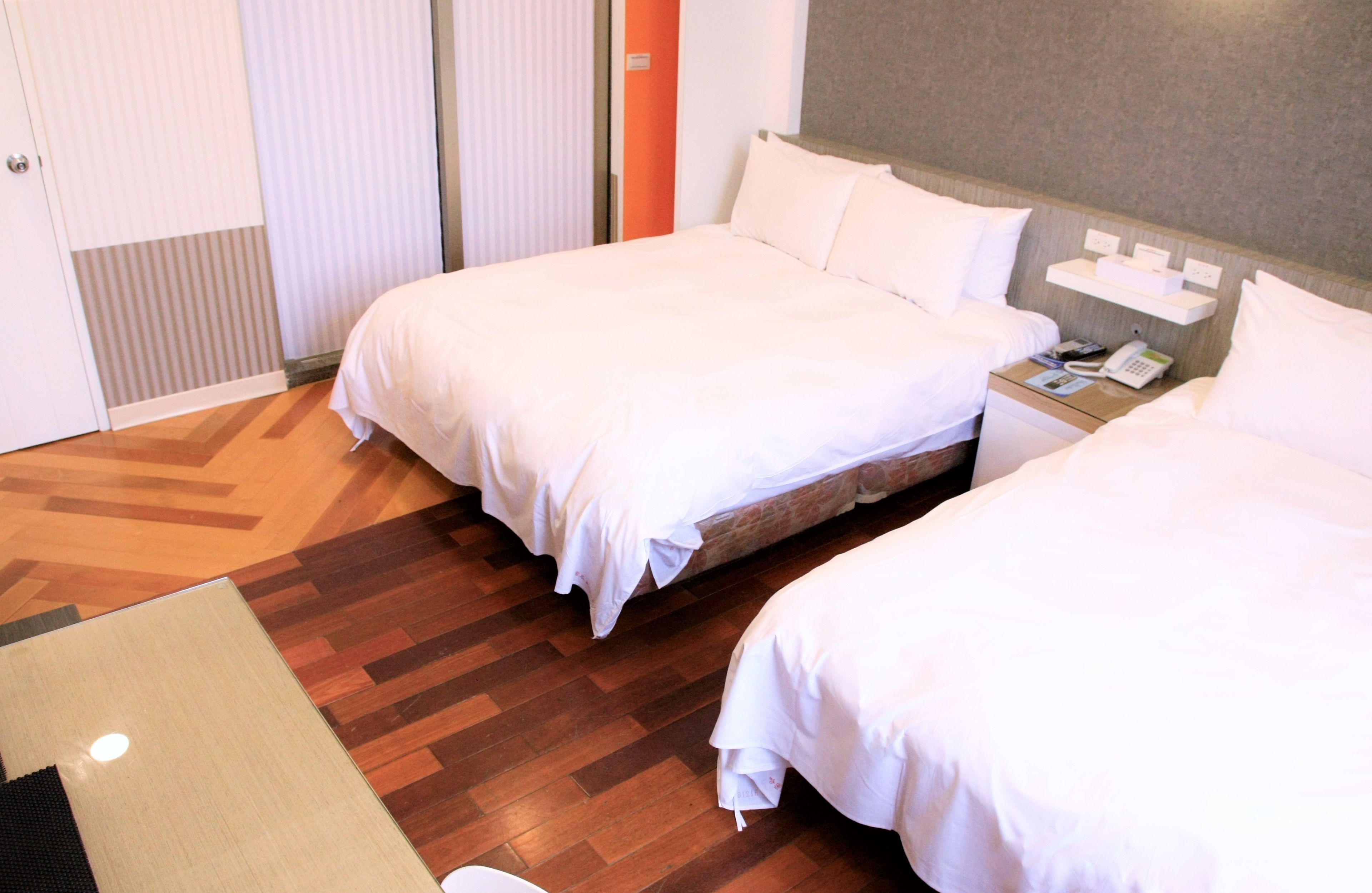 Quarto de hotel com duas camas, piso de madeira, mesa de cabeceira com telefone e revista, e prateleira na parede com produtos de higiene pessoal.