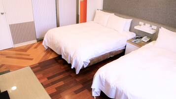 Quarto de hotel com duas camas, piso de madeira, mesa de cabeceira com telefone e revista, e prateleira na parede com produtos de higiene pessoal.