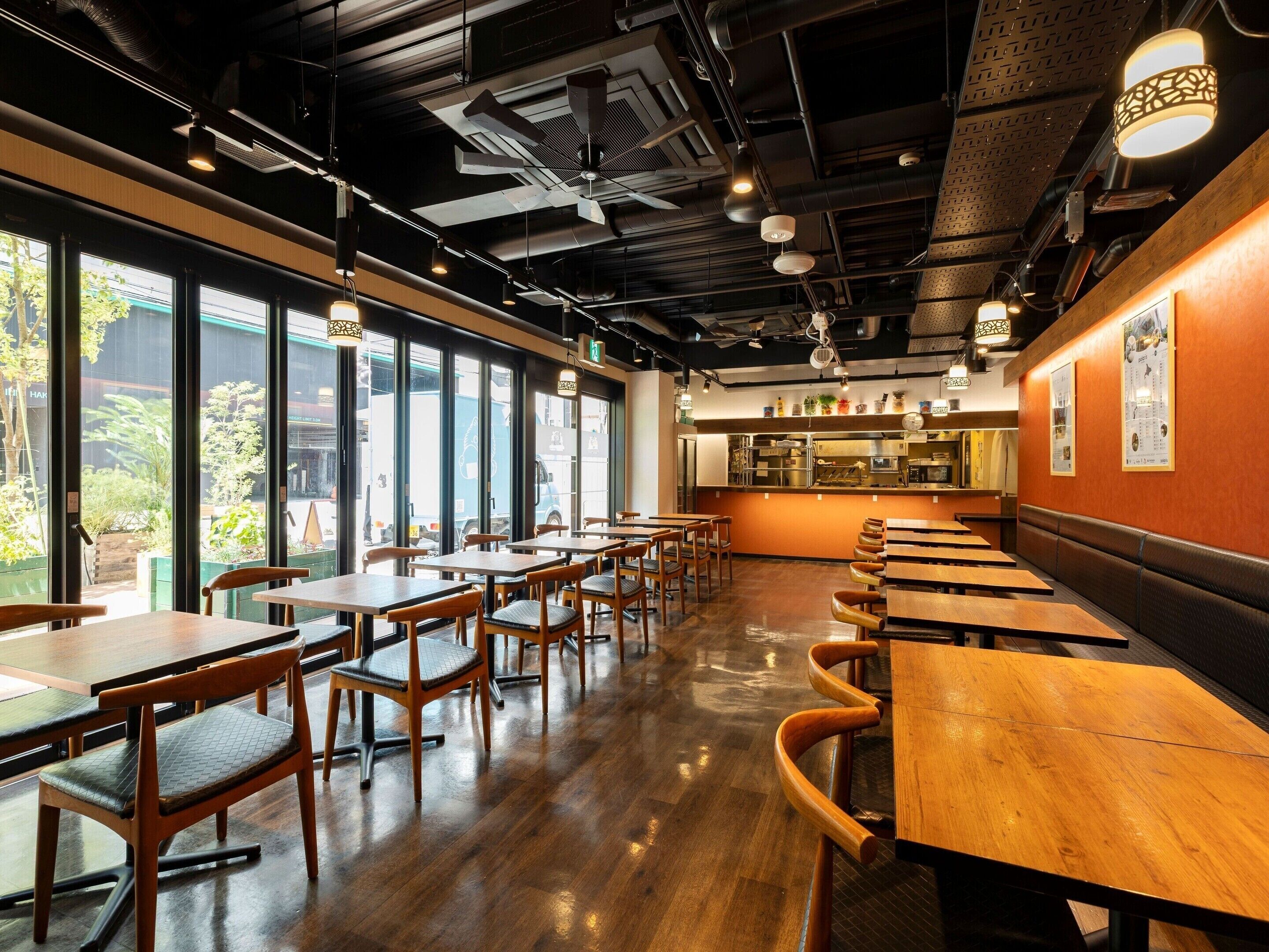 Interior de restaurante com mesas e cadeiras de madeira, área de cozinha e grandes janelas.