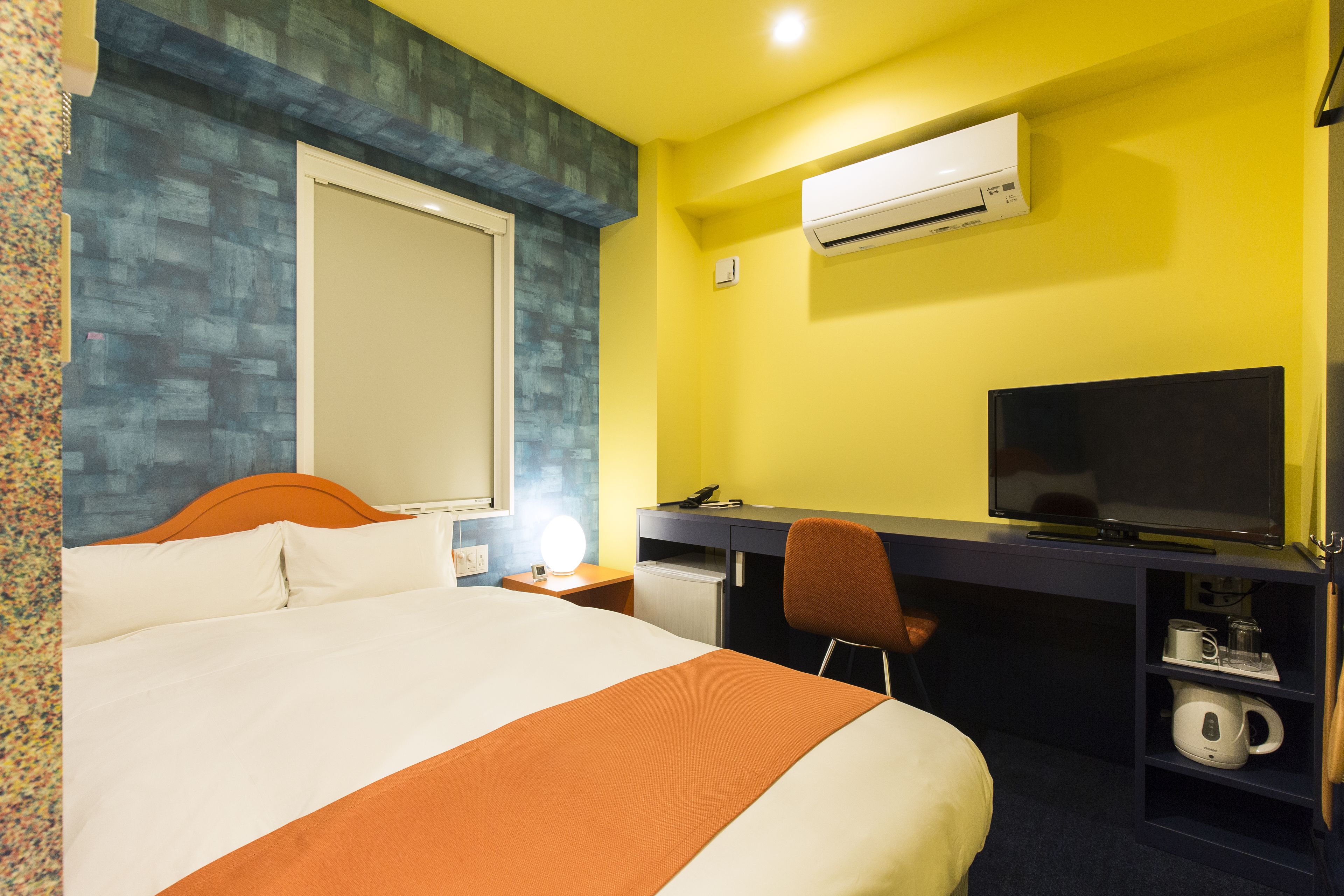 Quarto de hotel com cama, secretária com computador e televisão, ar condicionado e uma pequena cozinha.