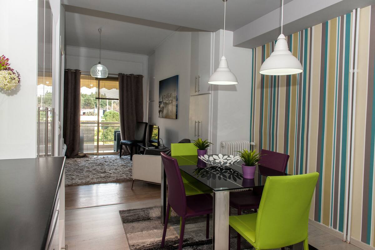 Uma área de refeições moderna com mesa de vidro, cadeiras verdes e roxas, e papel de parede listrado.