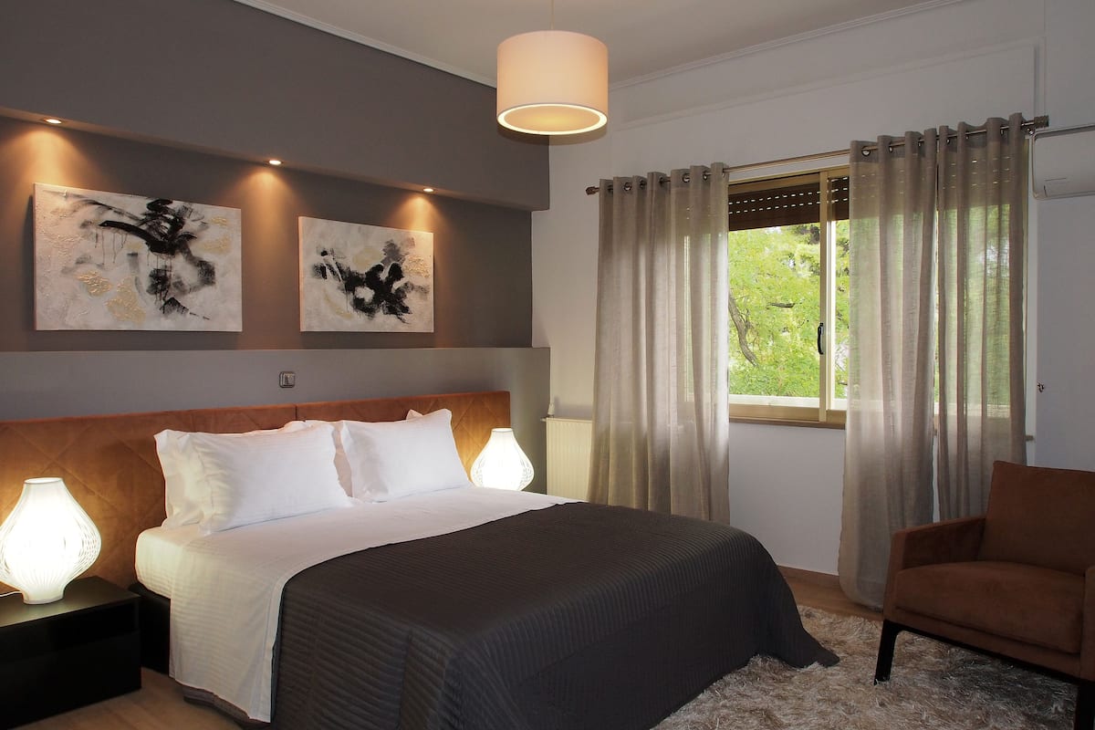 Quarto moderno com uma cama grande, duas pinturas penduradas na parede, uma luminária no teto, uma janela com cortinas e uma mesa de cabeceira com abajur.
