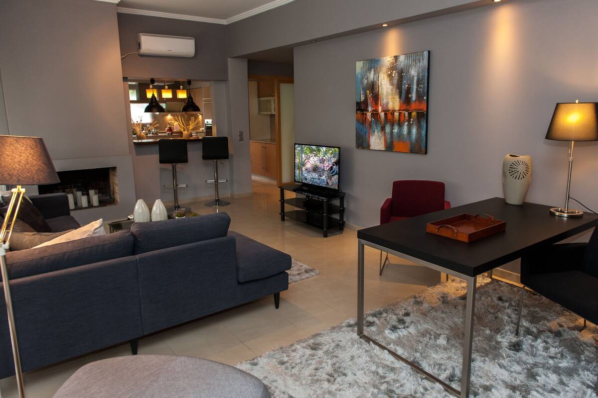 Sala moderna com sofá, TV e mesa de jantar. Cozinha visível ao fundo.