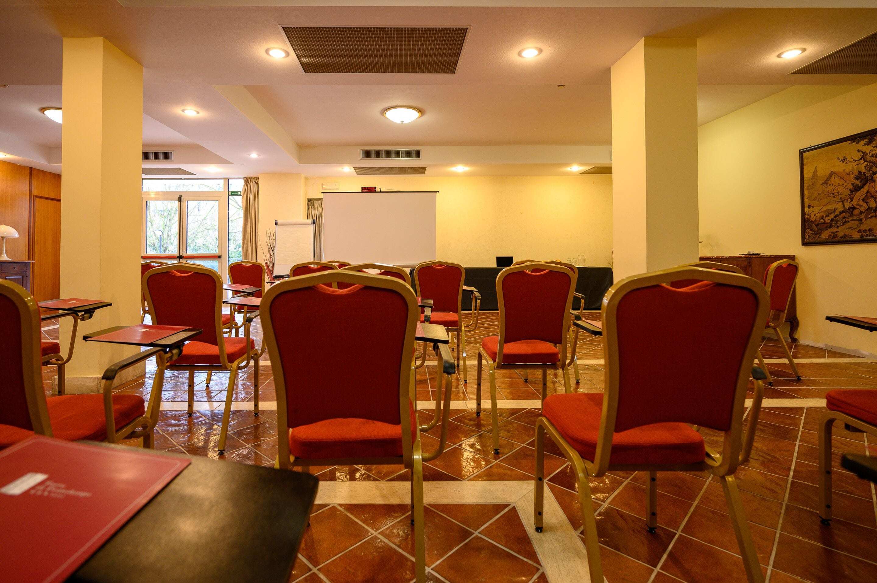 Uma sala de conferência com cadeiras e mesas vermelhas, um quadro branco e uma pintura na parede.