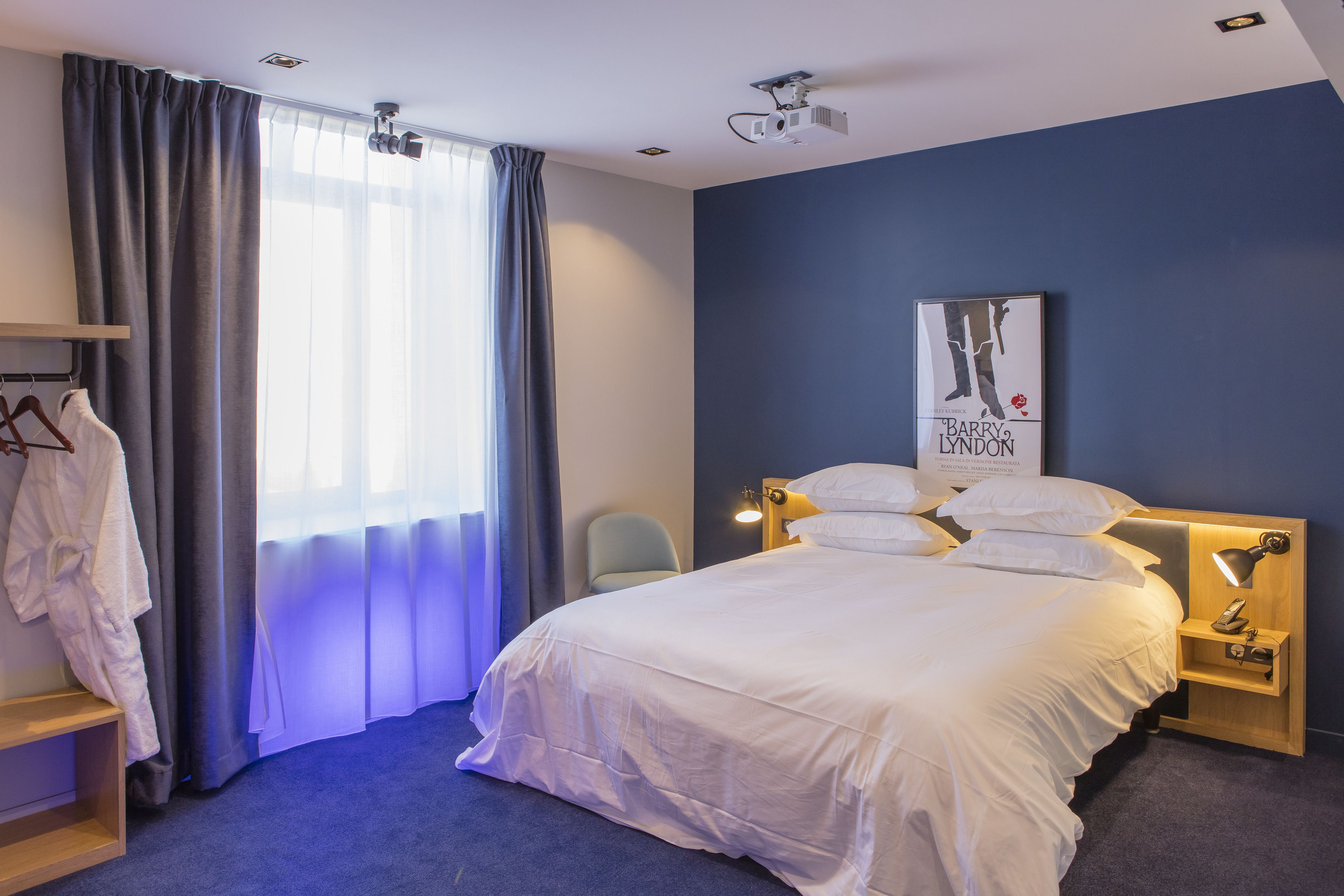 Quarto de hotel com uma cama grande, mesa de cabeceira com abajur, uma cadeira, guarda-roupa e janela com cortinas.