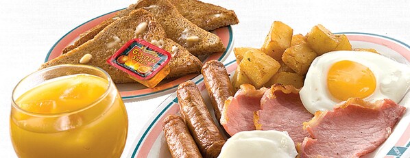 Prato de café da manhã com torradas, ovos, bacon, linguiça, batata palha e um copo de suco de laranja.