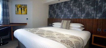 Quarto de hotel com cama, escrivaninha, cadeira e parede com padrão de árvore.