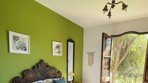 Quarto com parede verde, cabeceira de madeira, colcha florida, um espelho grande e vista para uma varanda com uma árvore.