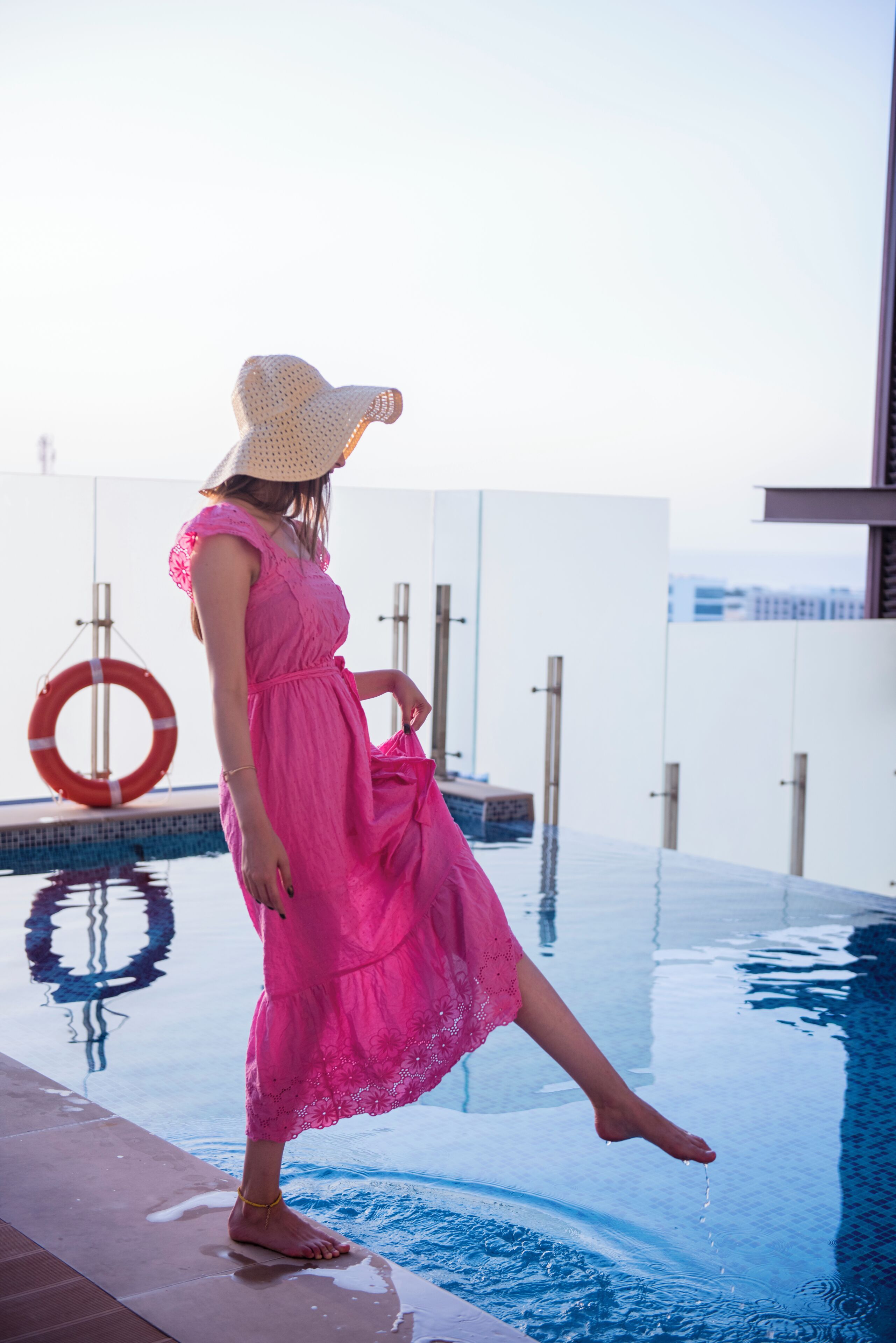 Mulher de vestido rosa e chapéu de palha junto a uma piscina, com um salva-vidas e um edifício ao fundo.