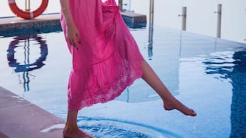 Mulher de vestido rosa e chapéu de palha junto a uma piscina, com um salva-vidas e um edifício ao fundo.