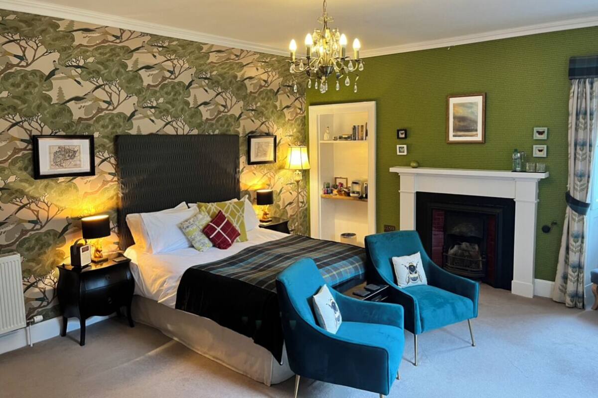 Quarto com cama, duas poltronas azuis, um candelabro, lareira e uma estante de livros.