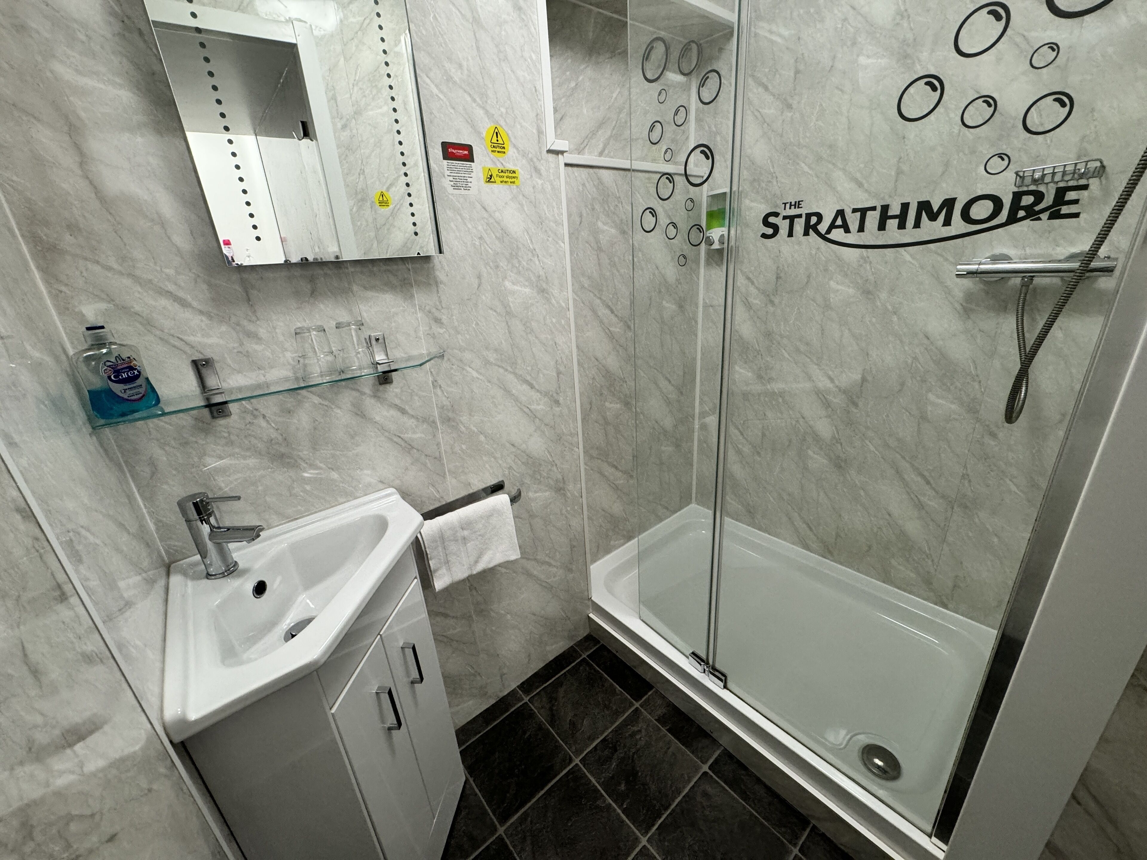 Casa de banho moderna com cabine de chuveiro em vidro, lavatório branco com espelho em cima e estante de vidro com produtos de higiene.