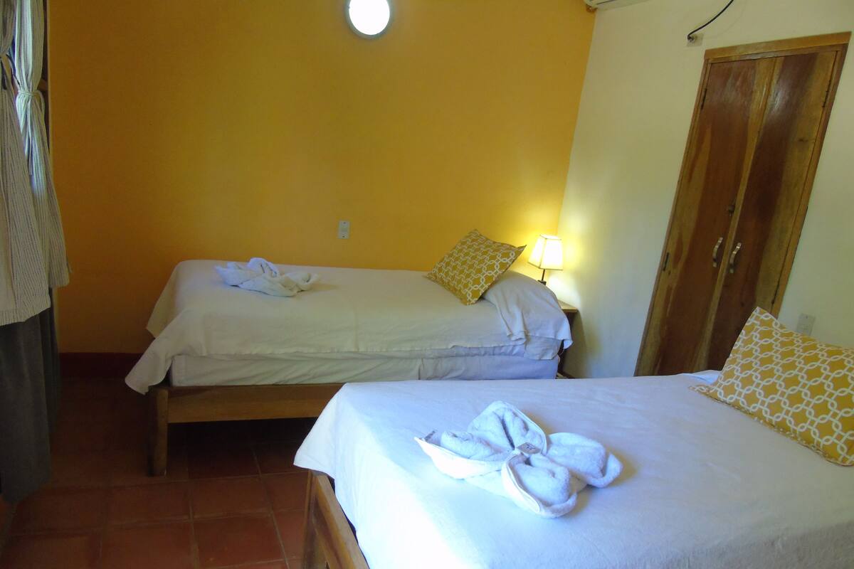 Quarto de hotel com duas camas, cada uma com roupa de cama e toalhas brancas, um guarda-roupa de madeira e uma parede amarela.