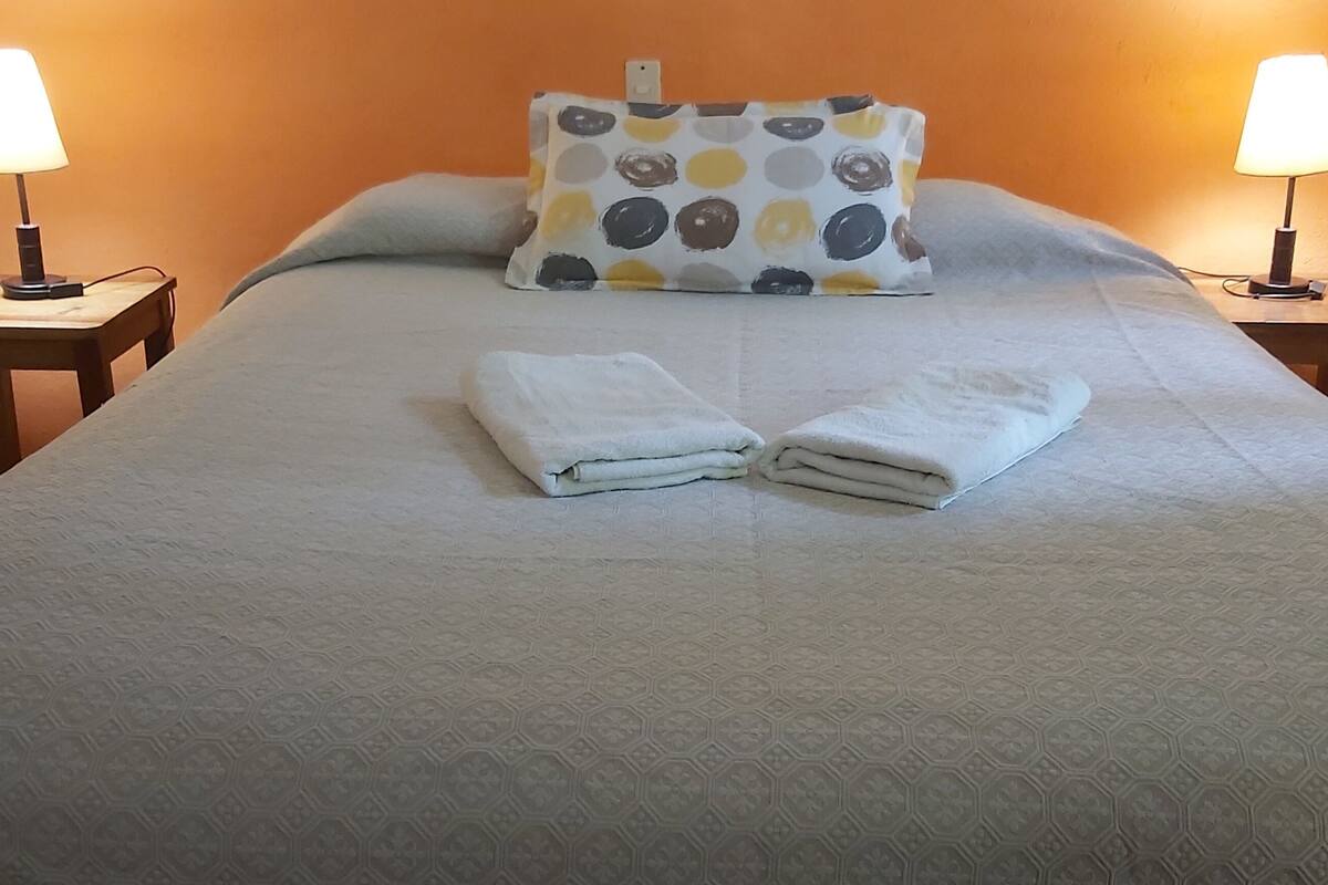 Cama bem arrumada com duas toalhas dobradas, um travesseiro estampado e duas luminárias em mesas de cabeceira de madeira.