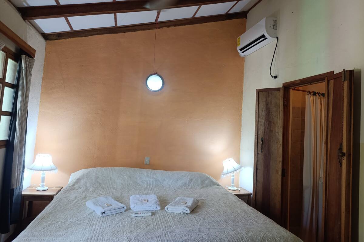 Quarto de hotel com cama, duas luminárias de cabeceira, aparelho de ar condicionado, guarda-roupa de madeira e ventoinha no teto.