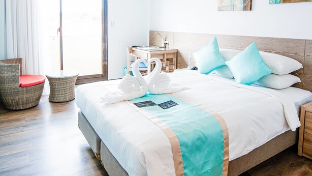 Quarto de hotel com uma cama grande, piso de madeira, uma mesinha, uma cadeira e uma janela com cortinas.