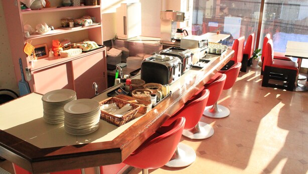Uma mesa de pequeno-almoço com cadeiras vermelhas, uma torradeira e diversas opções para o pequeno-almoço.