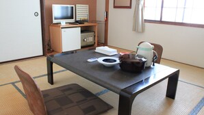 Um quarto japonês tradicional com tatames, uma mesa baixa, um banco acolchoado, uma televisão, um pequeno frigorífico e uma janela com cortinas.