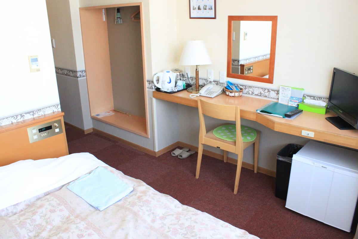 Quarto de hotel com cama, escrivaninha, cadeira, abajur, telefone, televisão e um pequeno frigorífico.