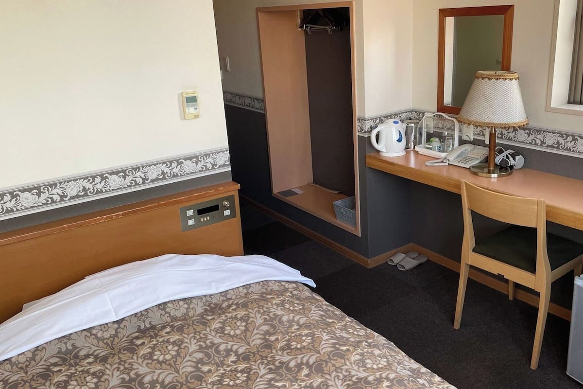 Quarto de hotel com cama, escrivaninha, espelho, abajur, telefone e pequeno frigorífico.