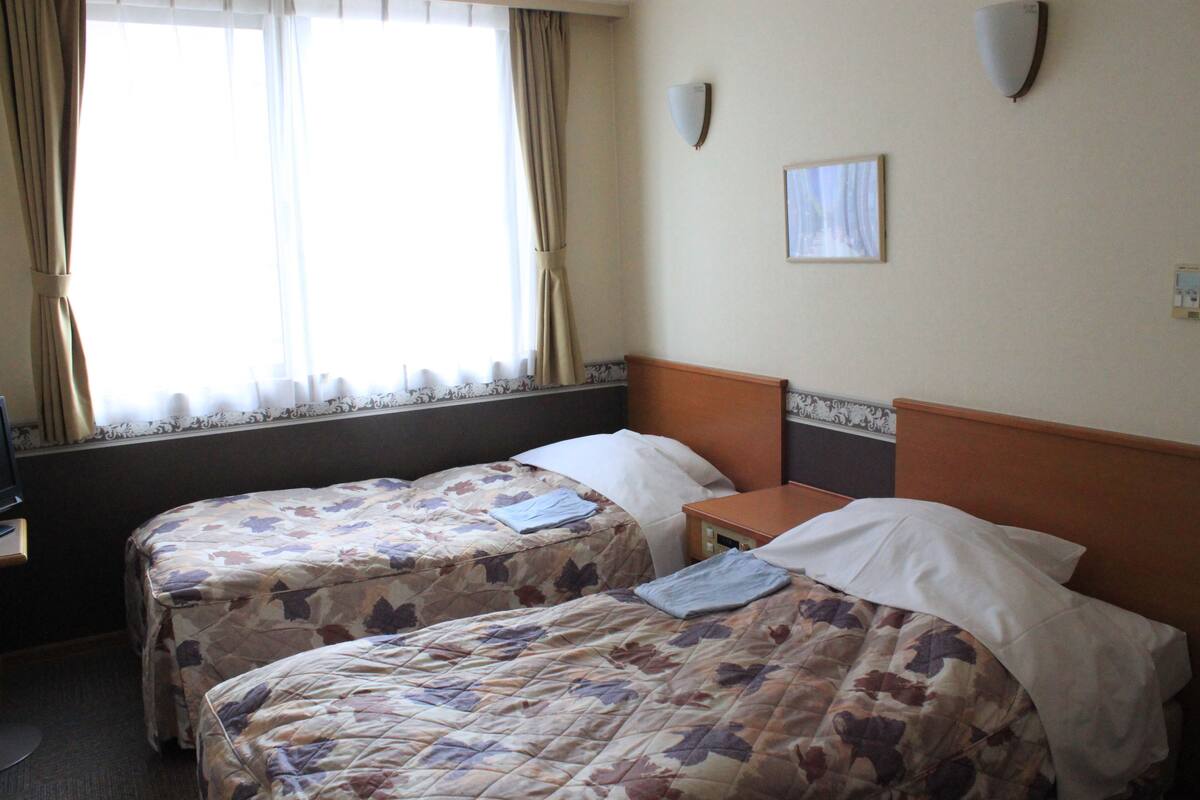 Quarto de hotel com duas camas, uma mesinha de cabeceira, televisão e janela com cortinas.