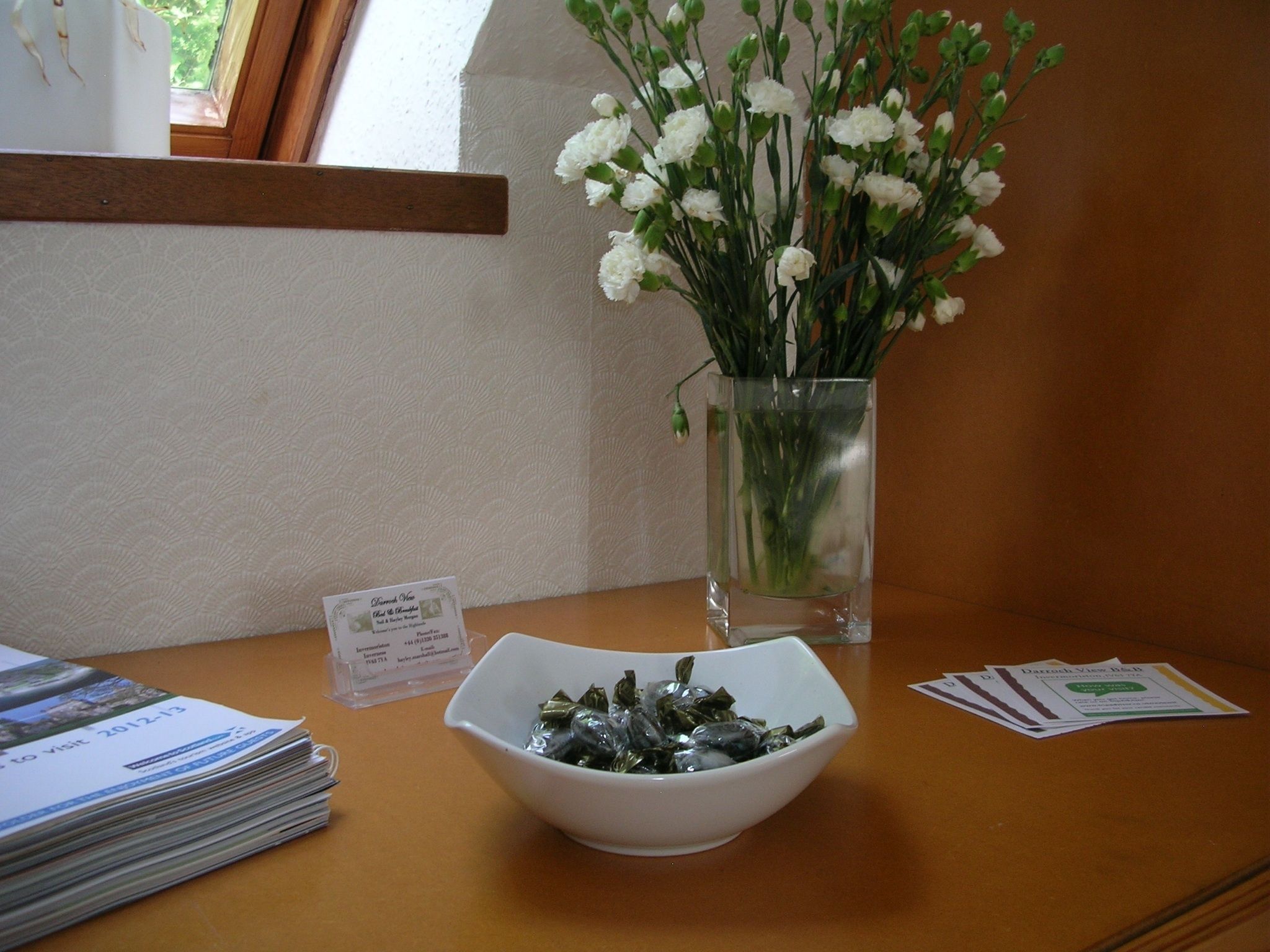 Uma mesa com um vaso branco contendo flores brancas, uma tigela branca com folhas verdes e uma pilha de folhetos.