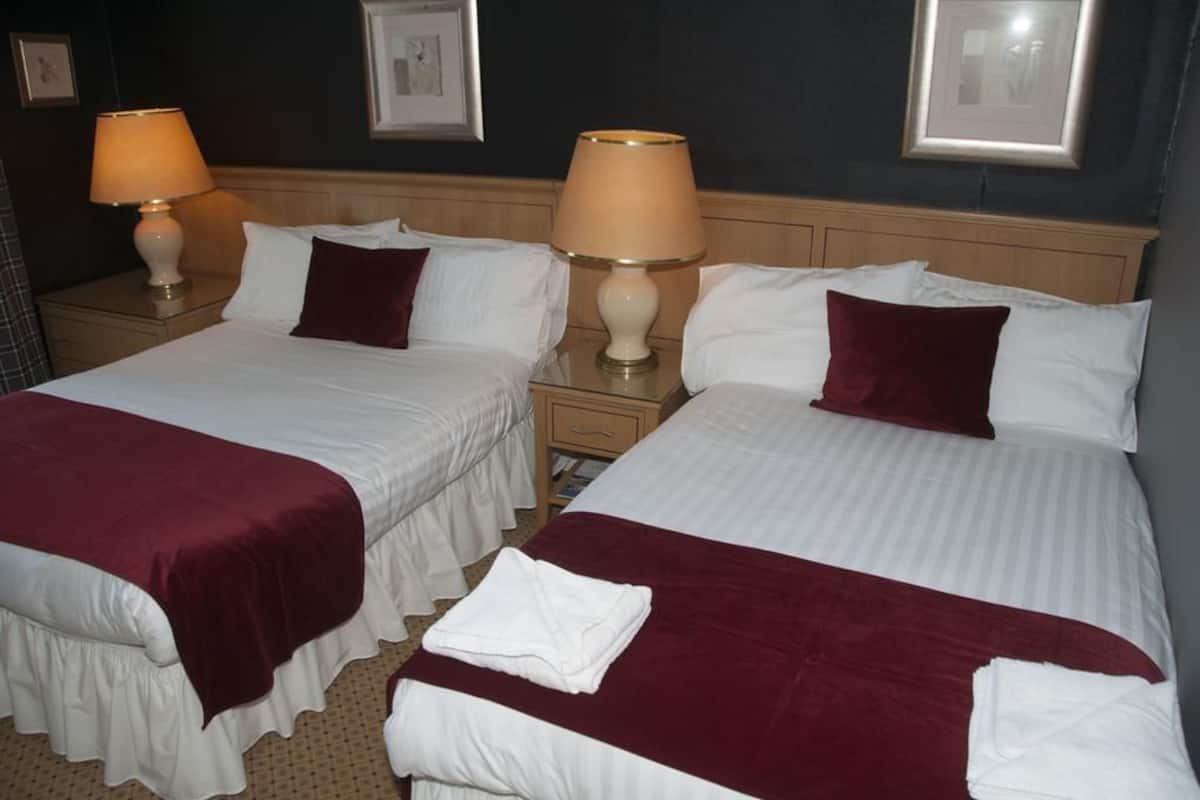 Quarto de hotel com duas camas, cada uma com roupa de cama branca e um cobertor vermelho. Há duas luminárias de cabeceira em mesas de cabeceira de madeira.