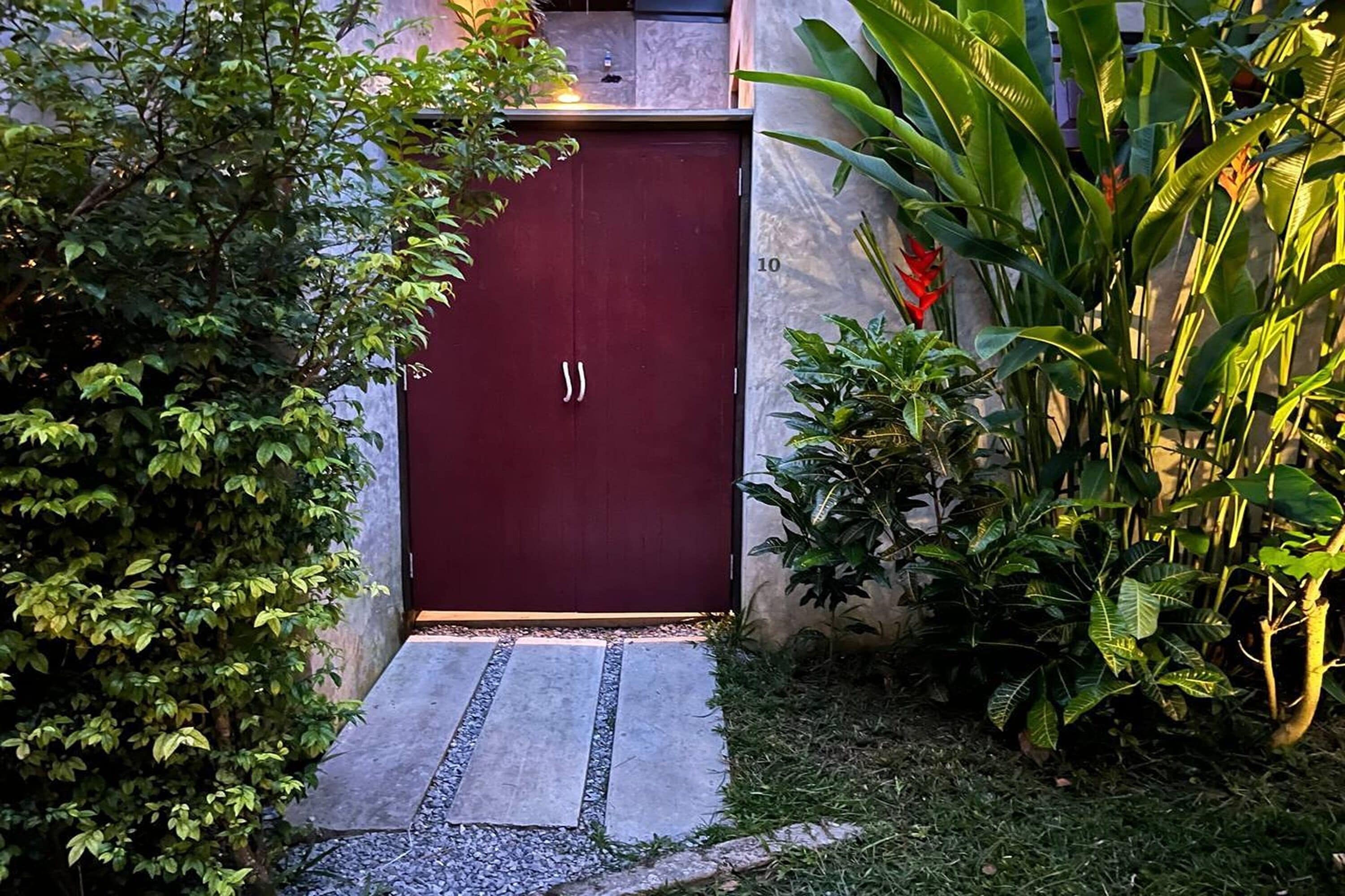 Uma porta vermelha com um pequeno degrau que a antecede, cercada por vegetação exuberante e um caminho de pedra.