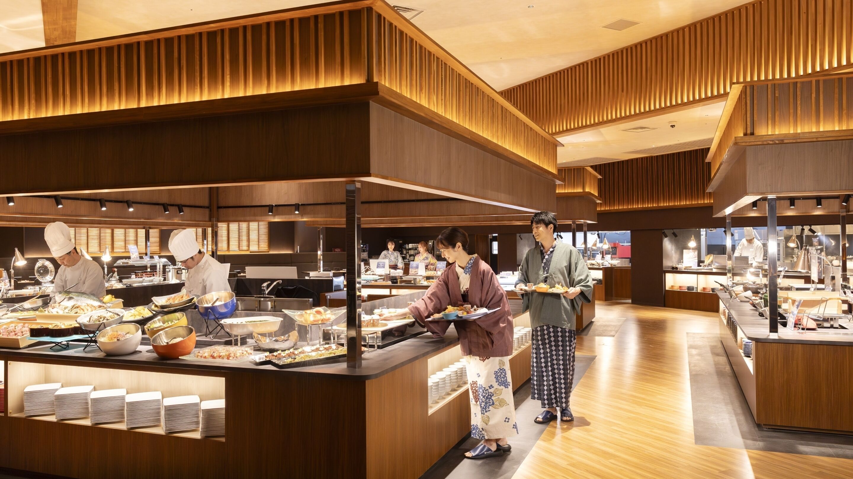 Disposição de buffet com diversas estações de serviço, variedade de alimentos e funcionários atendendo os hóspedes.