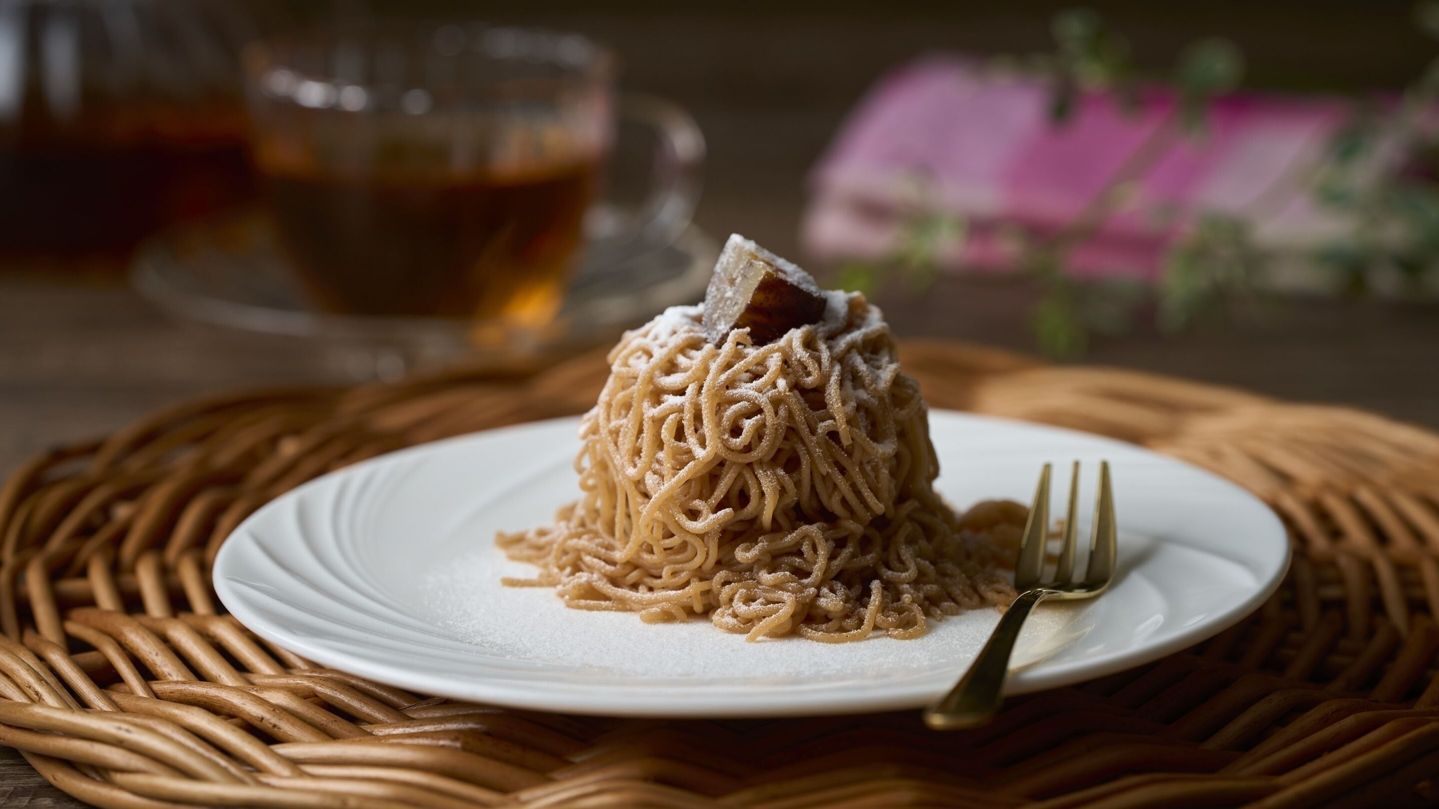Um prato de macarrão com um pedaço de carne por cima, um garfo e uma xícara de chá ao fundo.