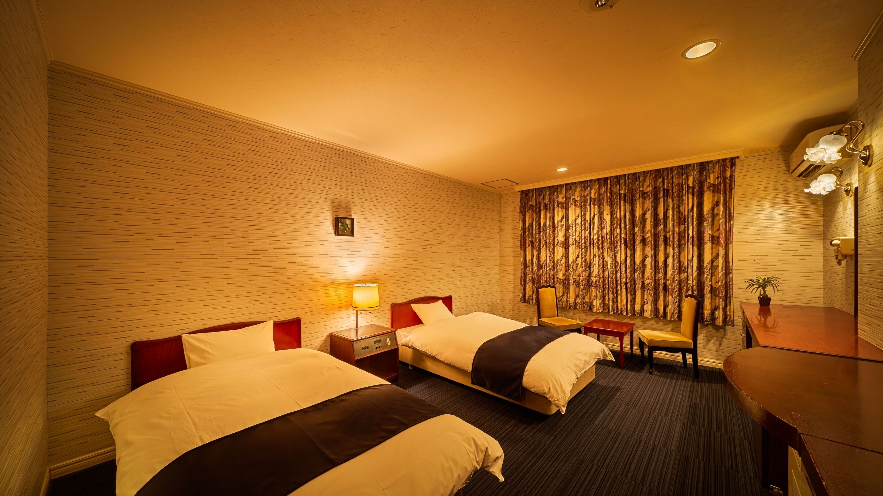 Quarto de hotel com duas camas, uma escrivaninha, uma cadeira, uma mesinha e uma janela grande com cortinas.