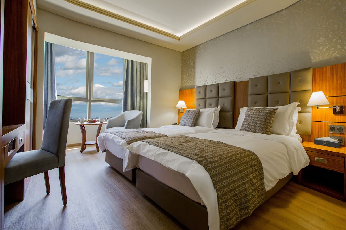 Quarto de hotel com uma cama grande, duas mesas de cabeceira, uma cadeira, uma escrivaninha e vista do céu pela janela.