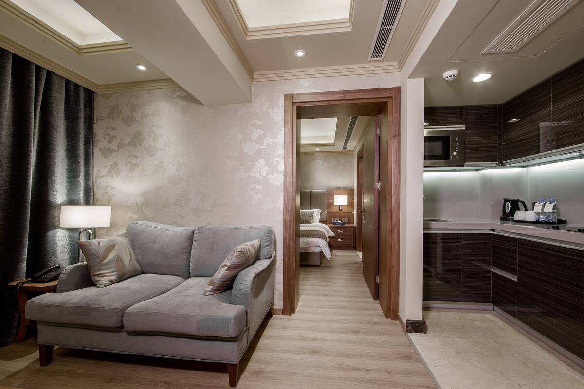 Quarto de hotel moderno com sofá, mesa de cabeceira com luminária, área de cozinha com micro-ondas e corredor que dá para outro quarto.