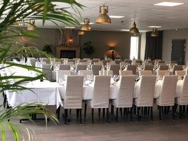 Uma sala de banquetes com várias mesas redondas preparadas para refeições, com toalhas brancas e cadeiras bege.