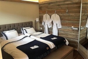 Uma cama de casal com cabeceira estofada, duas mesas de cabeceira e uma parede de madeira com um espelho e dois roués pendurados num suporte.