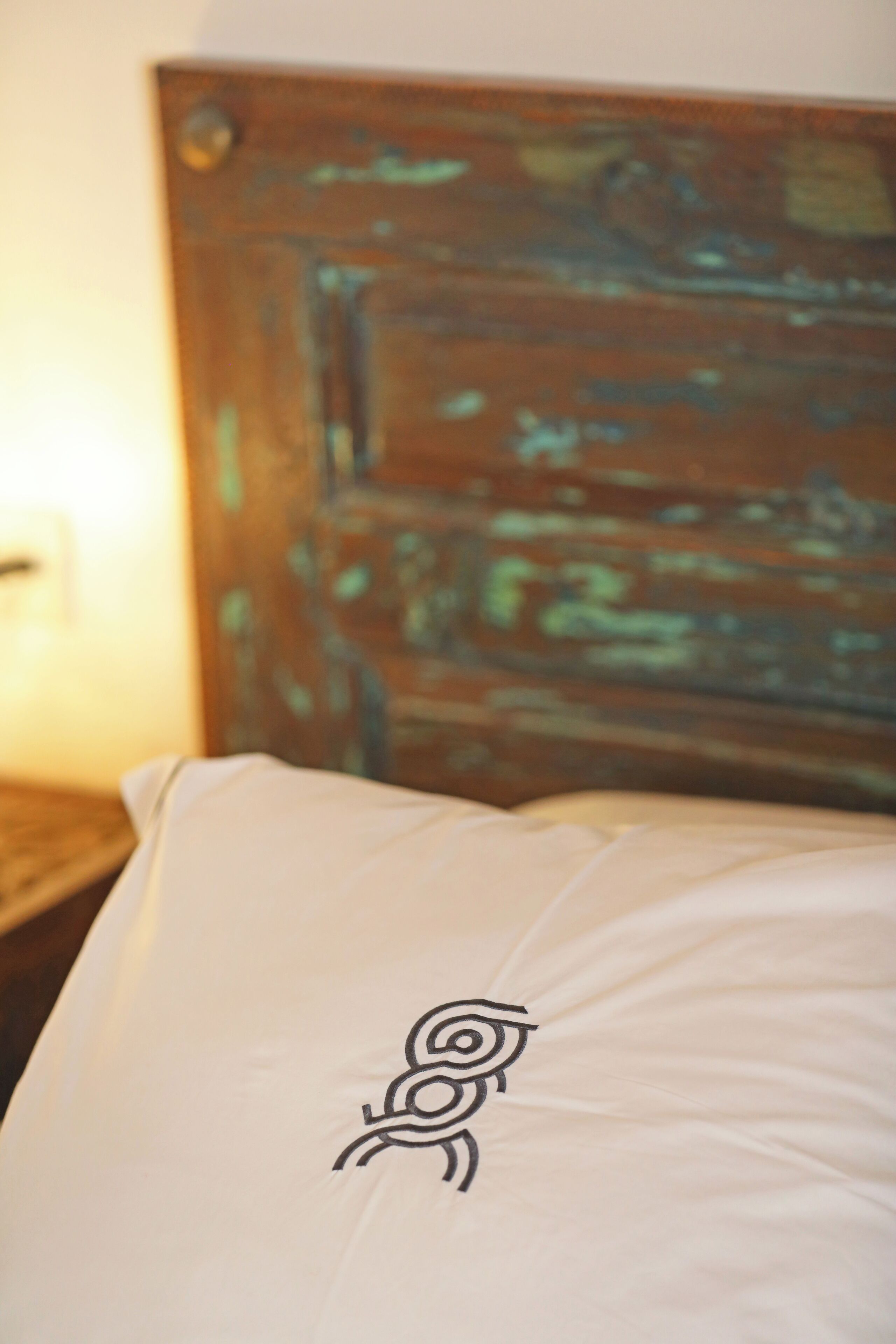 Cama com travesseiro branco e design preto, cabeceira de madeira e mesa de cabeceira de madeira com luminária.