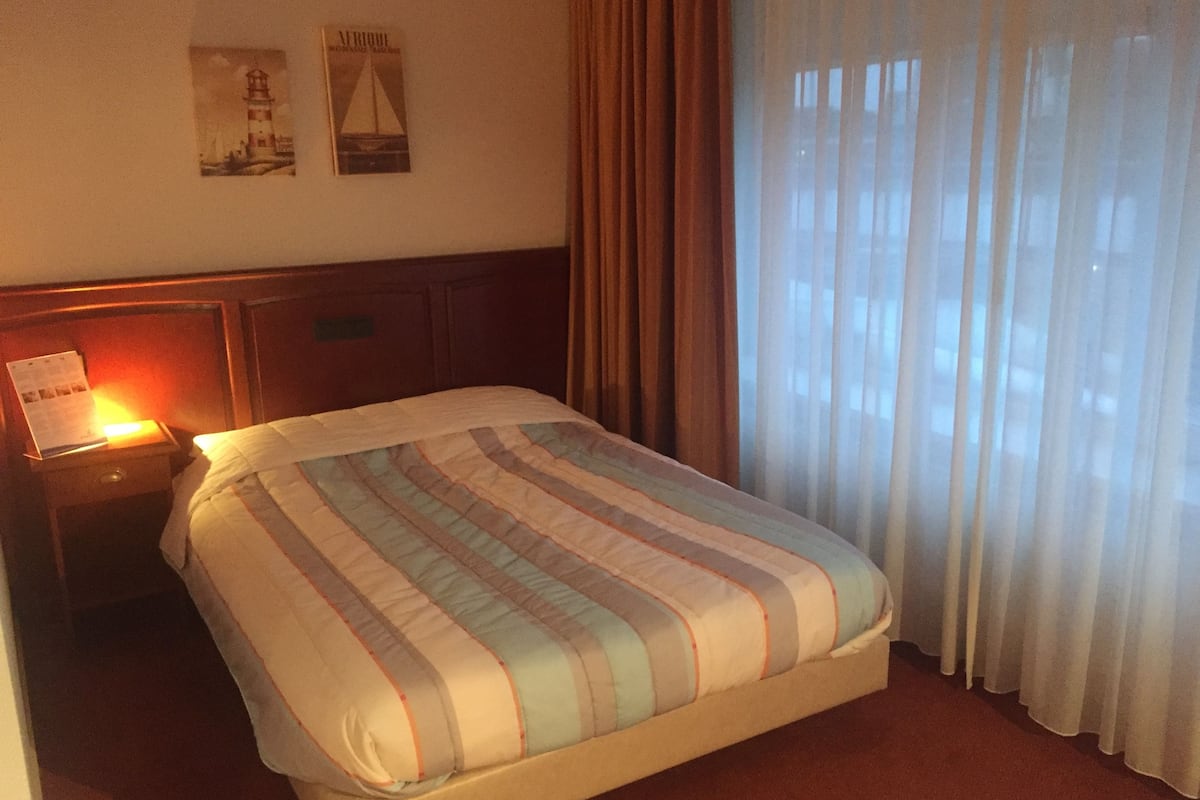 Quarto de hotel com cama, mesa de cabeceira, luminária, janela com cortinas e duas decorações na parede.