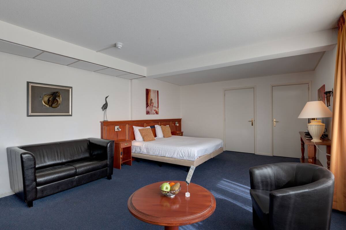 Quarto de hotel com cama, sofá de couro preto, mesa de centro de madeira com frutas e uma luminária.