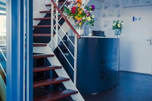 Escadaria com degraus de madeira e corrimão de metal, levando a uma mesa de recepção com um vaso de flores.