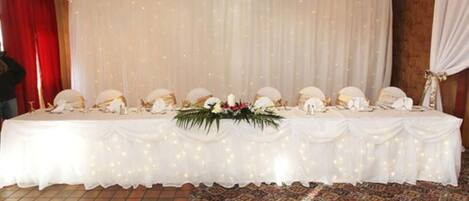 Uma mesa de banquete posta com toalhas brancas, cadeiras brancas e um arranjo floral.