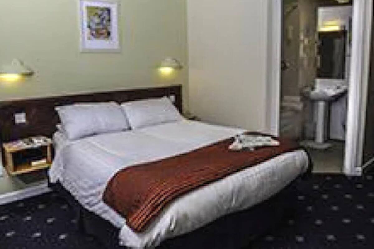 Quarto de hotel com cama, mesas de cabeceira, luminária, quadro na parede e banheiro visível por uma porta aberta.