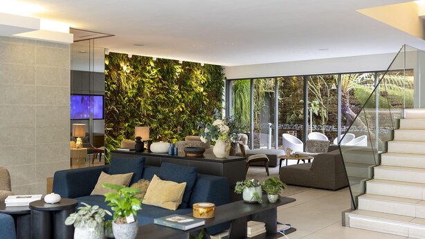 Uma sala de estar moderna com sofá, mesa de centro e plantas, uma TV e uma escada que leva a uma varanda com vista para o verde.