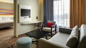 Quarto de hotel moderno com cama, televisão na parede, sofá, poltrona e uma janela grande com cortinas.