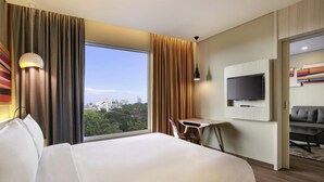 Quarto de hotel com uma cama grande, uma escrivaninha, uma cadeira, televisão na parede e vista para a paisagem urbana pela janela.