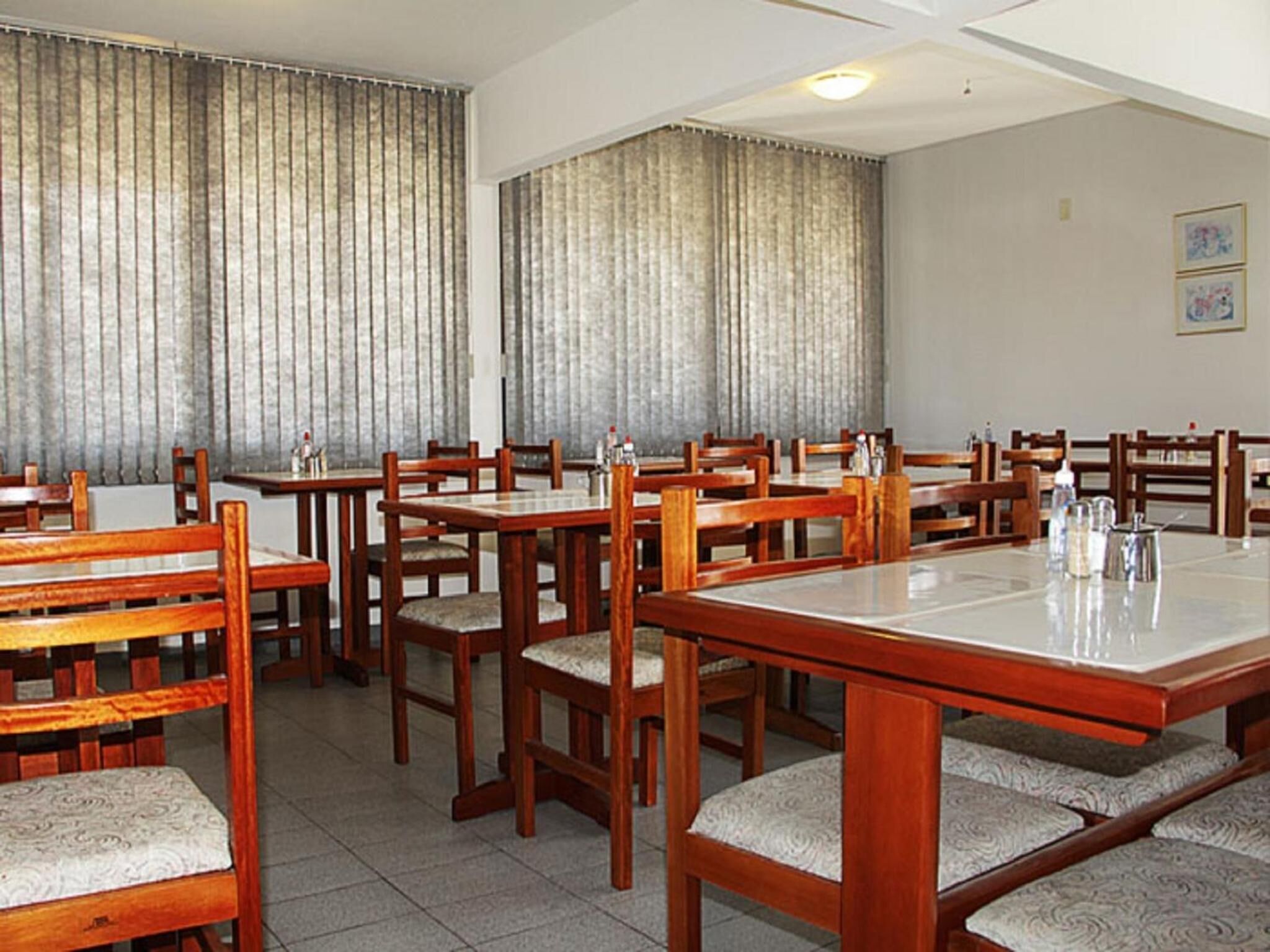 Uma sala de jantar com mesas e cadeiras de madeira, janelas amplas com cortinas e piso de azulejos.