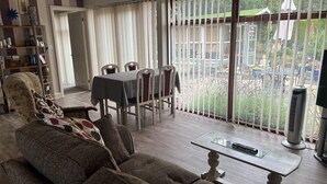 Uma sala de estar com sofá, mesa de jantar e cadeiras, televisão e uma janela grande com persianas.