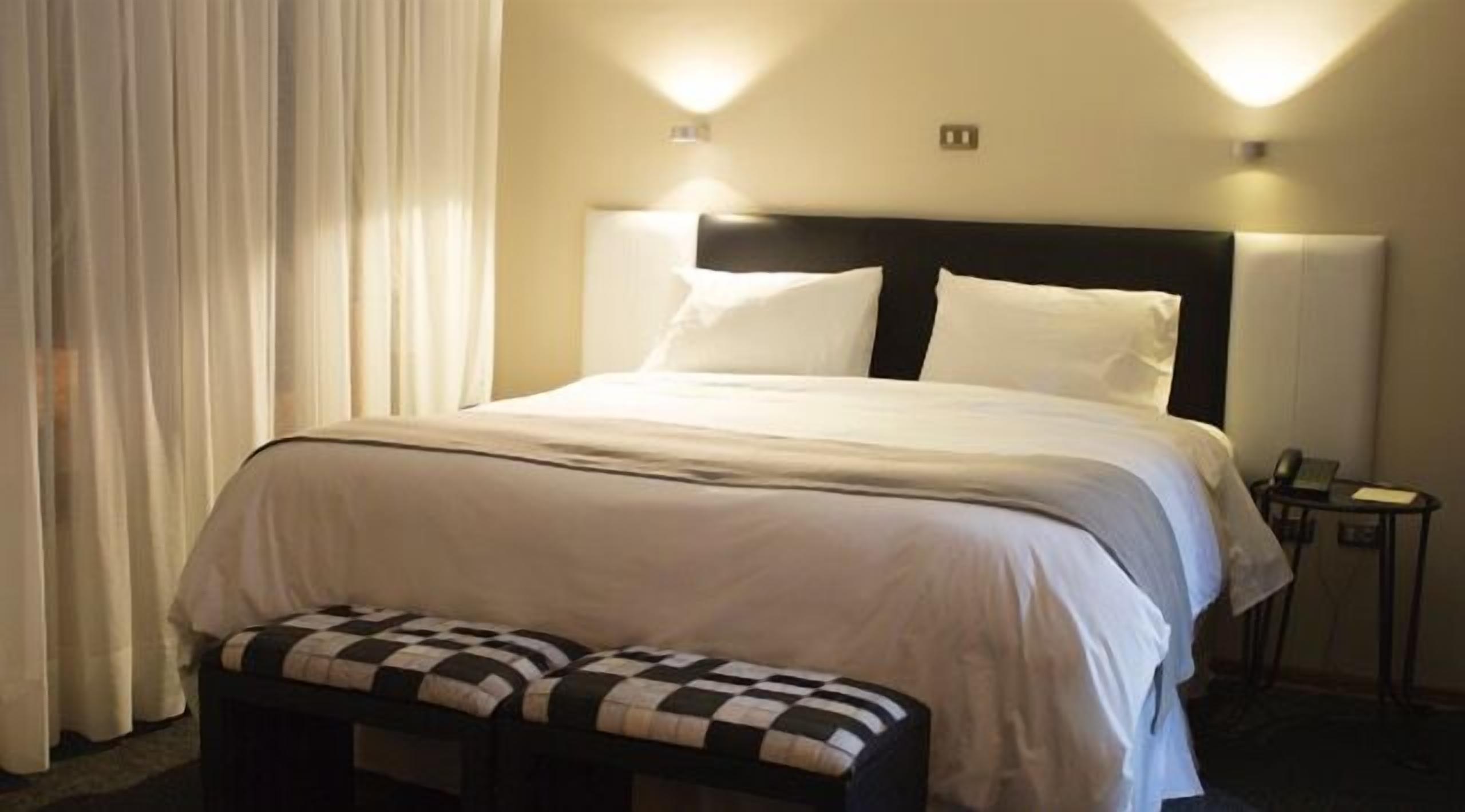 Quarto de hotel com uma cama grande, uma mesinha de cabeceira com telefone e um banquinho com almofada xadrez.