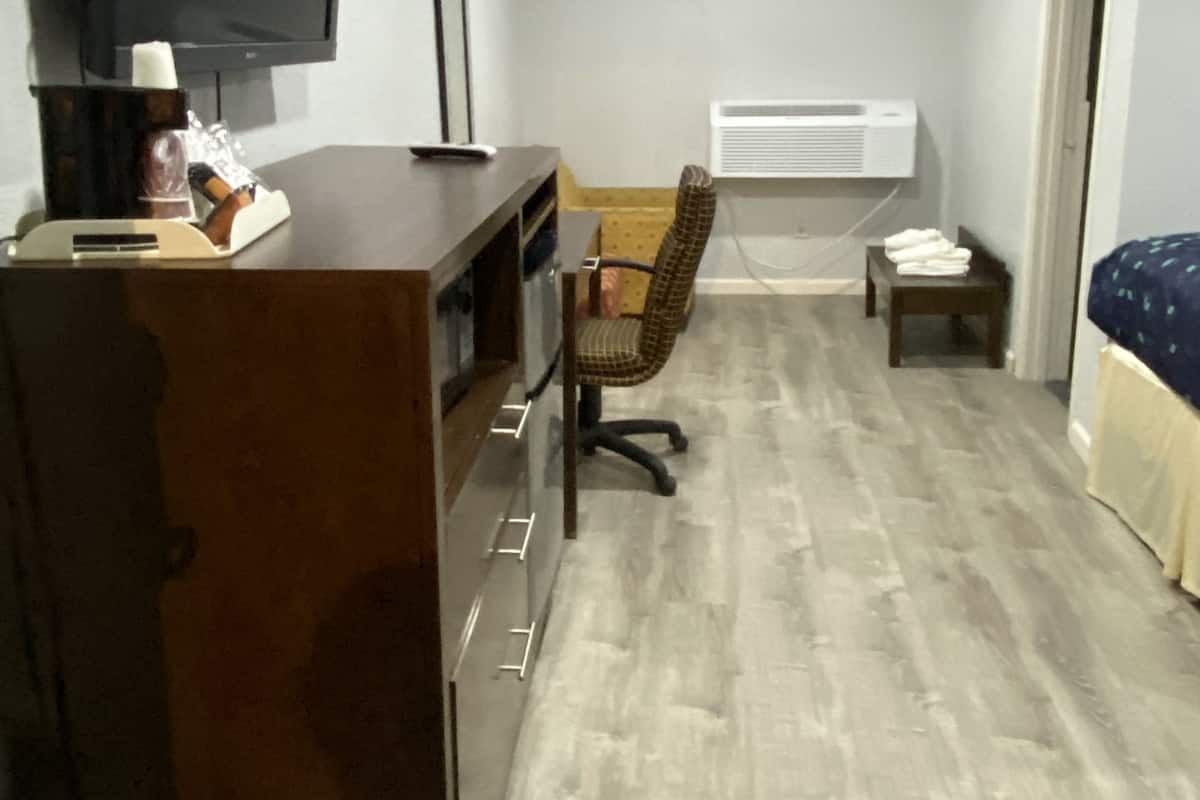 Quarto de hotel com escrivaninha de madeira, cadeira, cama, televisão, ar condicionado e pequena mesa com abajur.