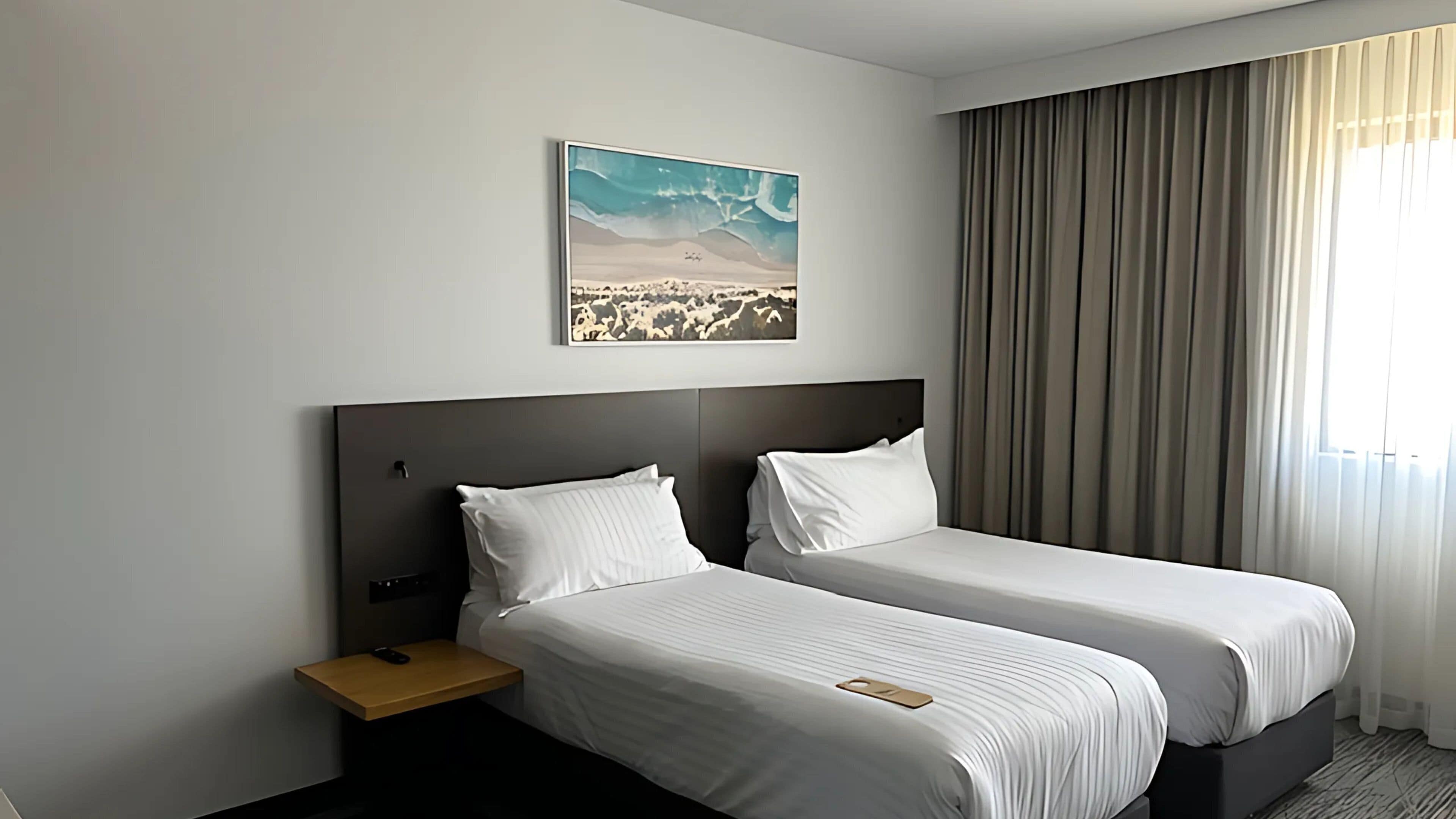 Quarto de hotel com duas camas, uma mesa de cabeceira, um controle remoto e uma pintura de uma paisagem desértica.